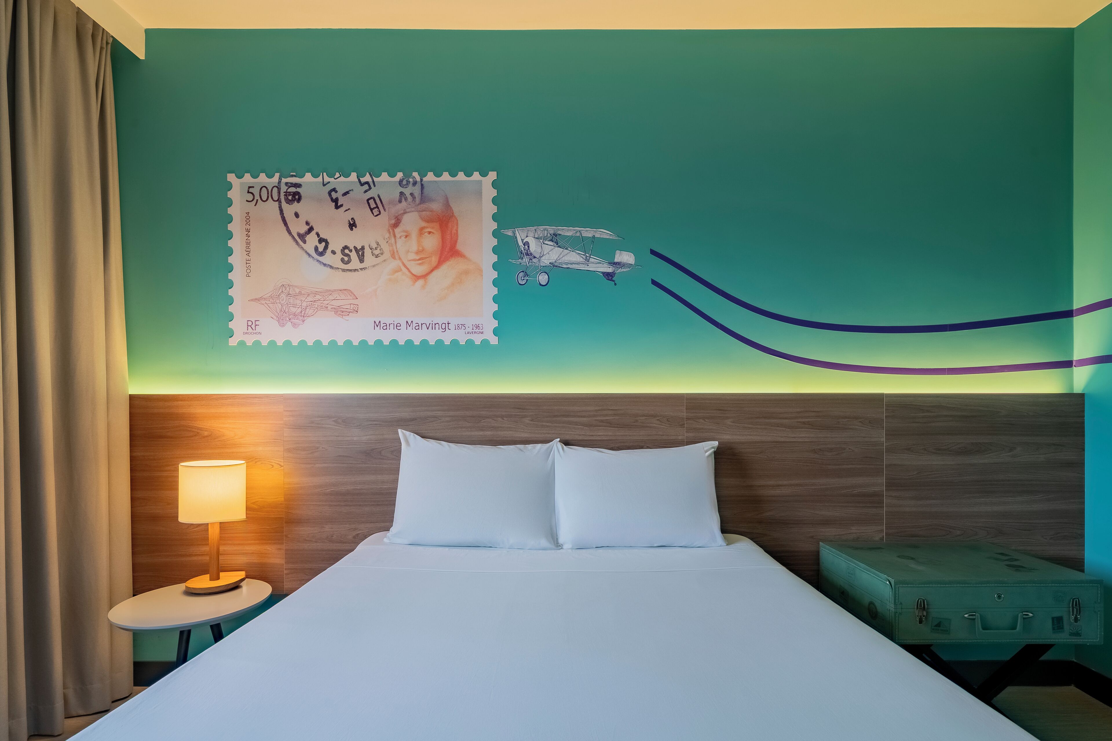 Quarto de hotel com cama, mesa de cabeceira, abajur, mala e selo na parede.