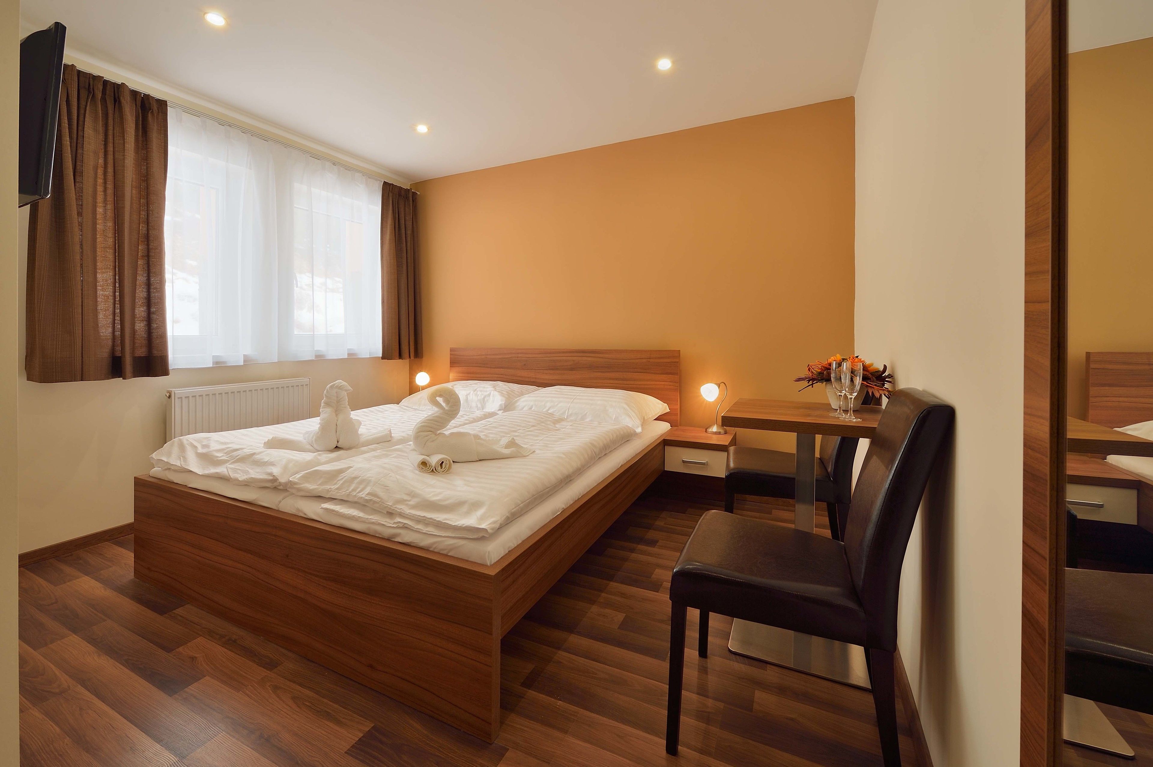 Quarto com cama de madeira, mesa de cabeceira com abajur, uma cadeira e uma pequena mesa com um vaso.