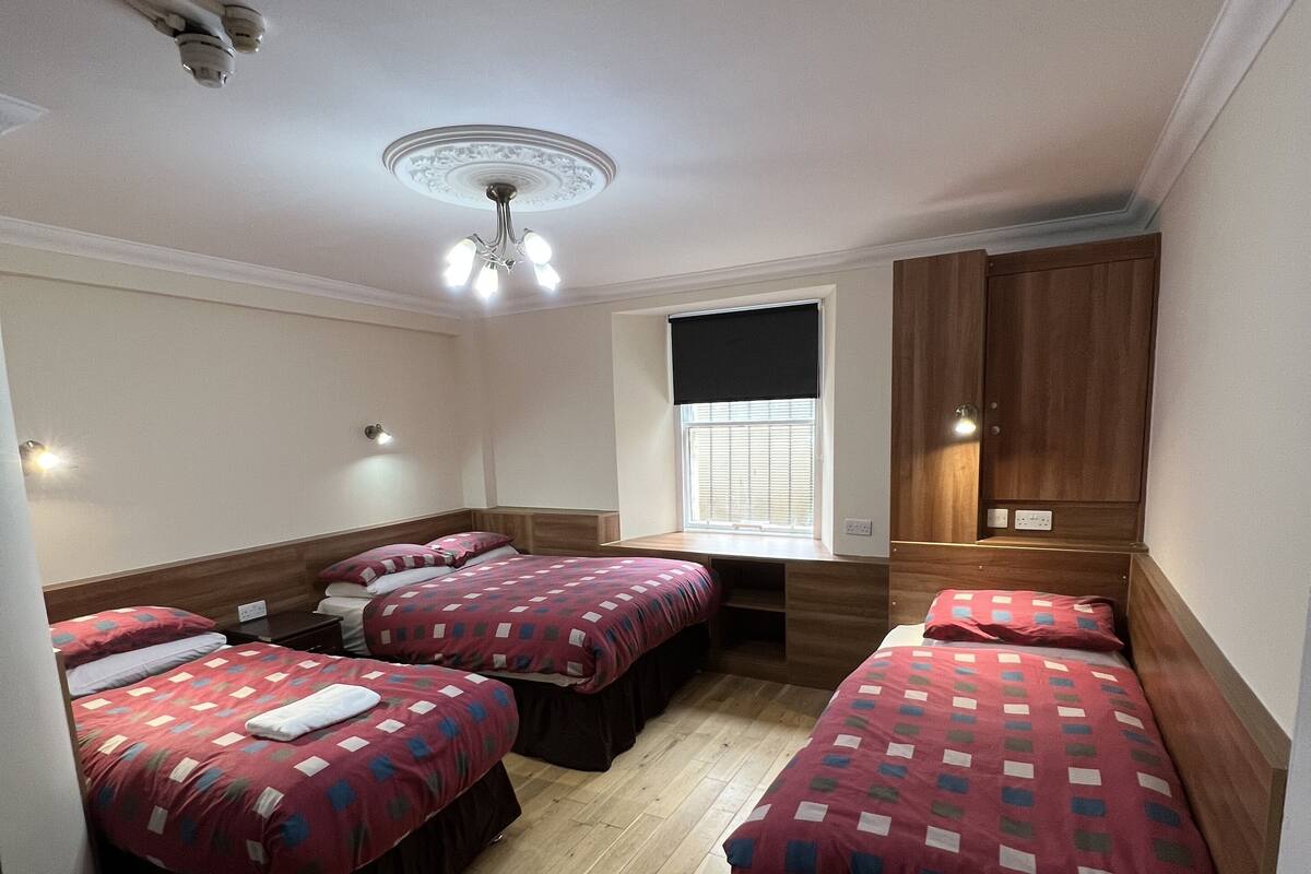 Quarto com quatro camas, piso de madeira, ventilador de teto e janela com persianas.