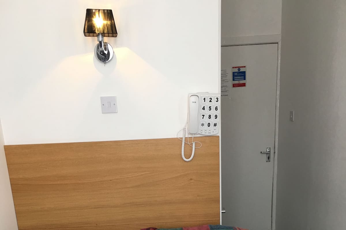 Quarto de hotel com cabeceira de madeira, edredom estampado, travesseiro branco e manta branca, além de uma toalha enrolada. Há um telefone fixado na parede e uma porta com uma placa.