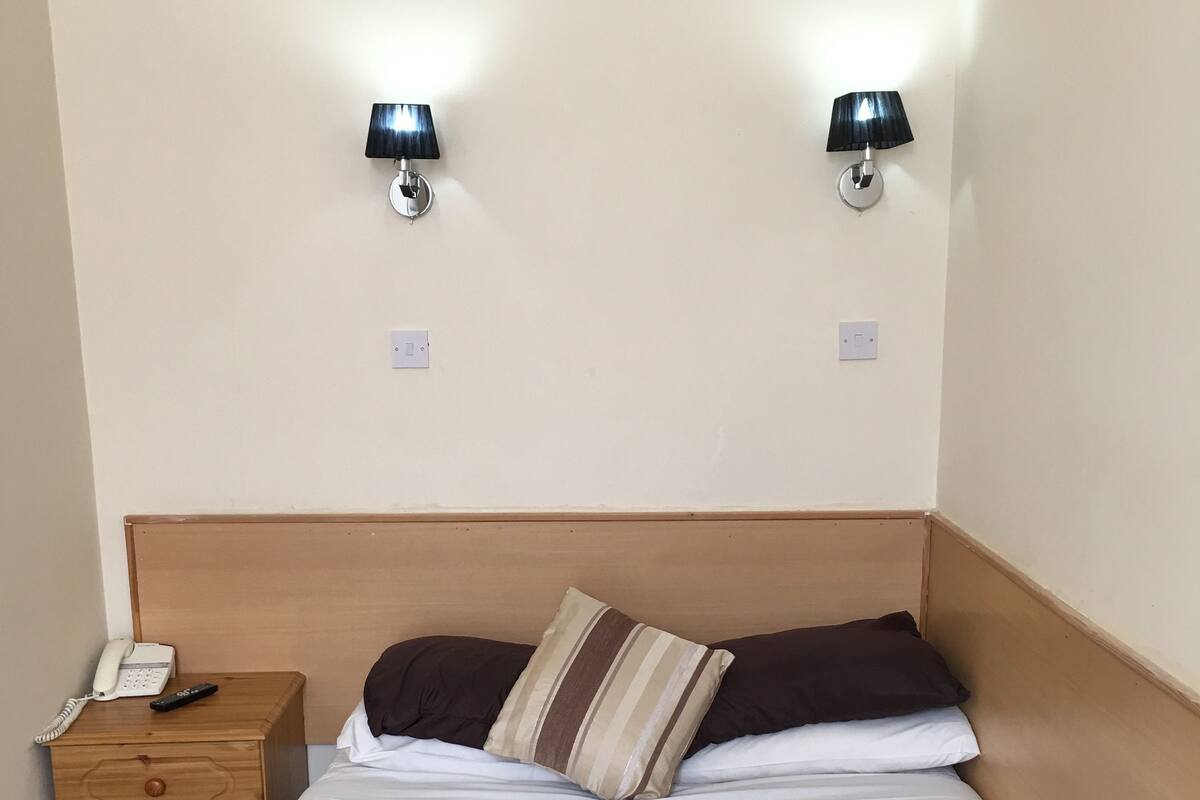 Cama bem arrumada com travesseiro listrado, duas luminárias de cabeceira, mesa de cabeceira de madeira com um telefone e um par de toalhas dobradas.