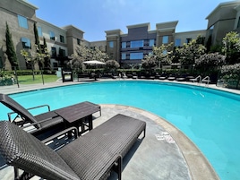 Piscina com espreguiçadeiras e uma mesa, rodeada por edifícios e vegetação.