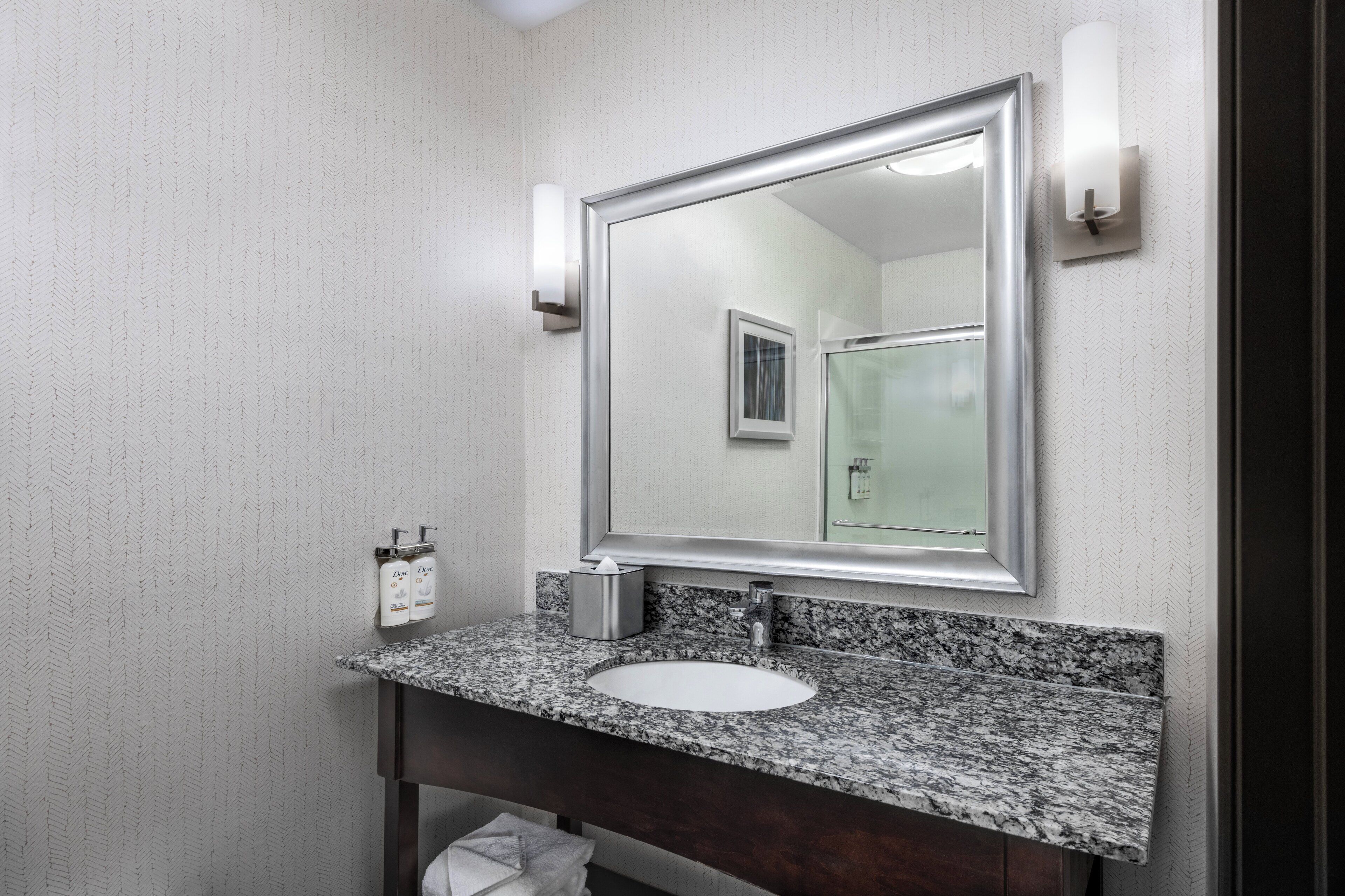 Casa de banho com bancada de granito, lavatório branco e espelho grande. Possui duas luminárias fixadas na parede e um dispensador de toalhas.