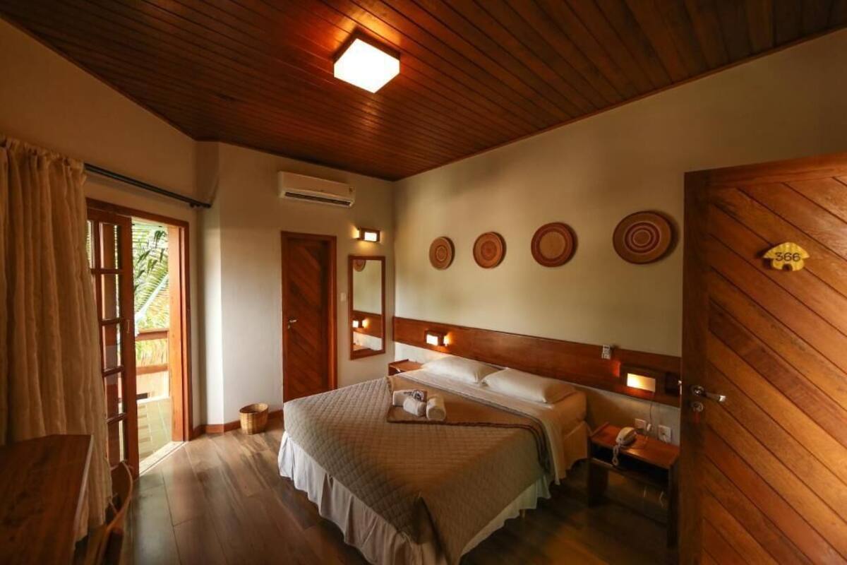 Quarto com painéis de madeira, cama, mesas de cabeceira, espelho e porta com placa de identificação.