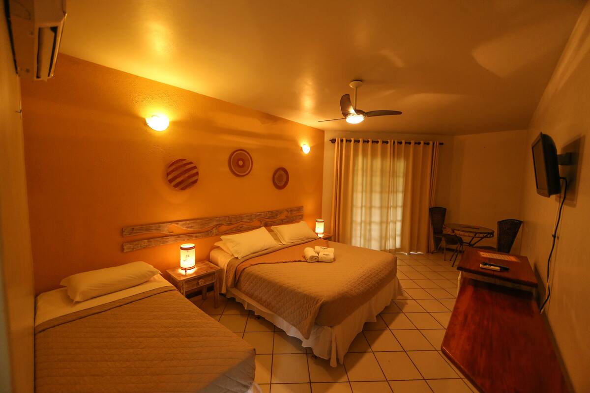 Quarto de hotel com duas camas, uma escrivaninha, uma cadeira, um ventilador de teto e uma televisão presa à parede.