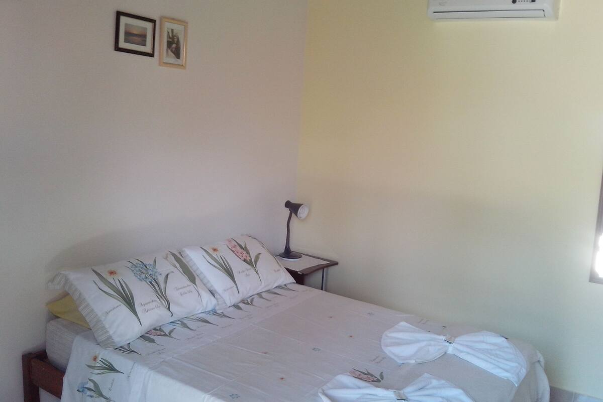 Uma cama bem arrumada com roupa de cama florida, uma luminária de cabeceira e duas fotos emolduradas na parede.