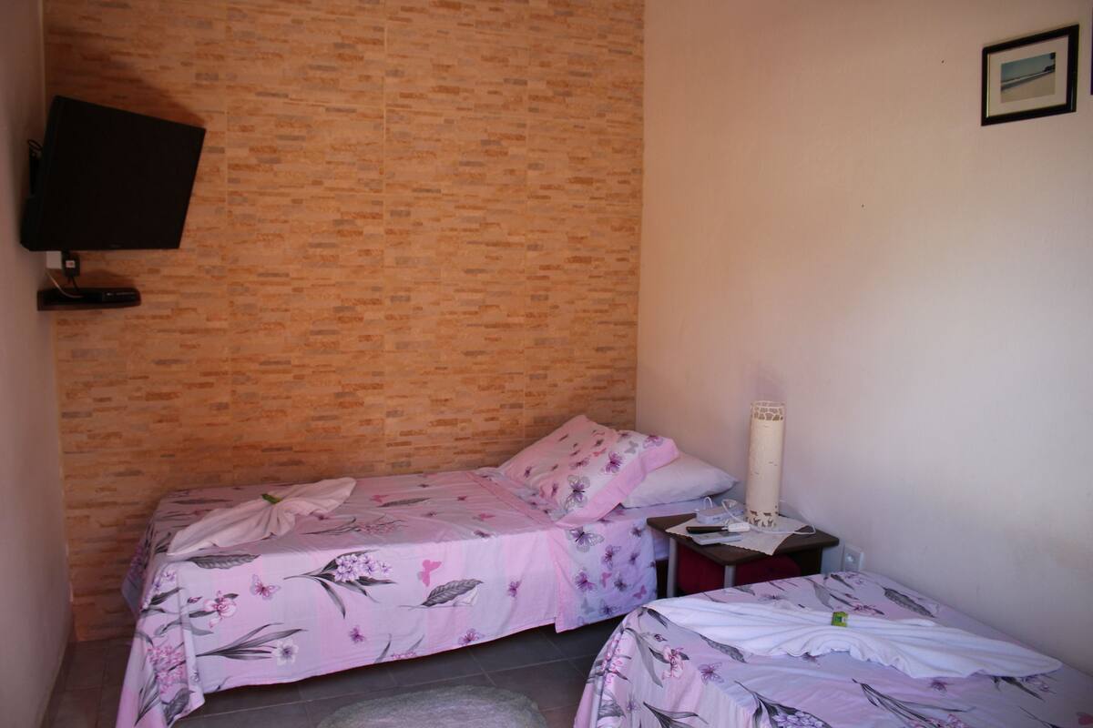 Quarto com duas camas, televisão de tela plana na parede e uma mesinha com abajur.