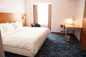 Quarto de hotel com uma cama grande, uma escrivaninha com cadeira, uma luminária e uma janela com cortinas.