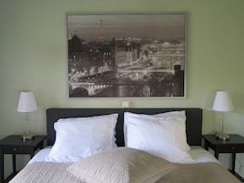 Quarto de hotel com uma cama, duas mesas de cabeceira e uma grande pintura em preto e branco de uma paisagem urbana na parede.