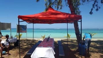 Um espaço de refeições à beira-mar com toldo vermelho, mesas preparadas para dois e vista para o oceano.