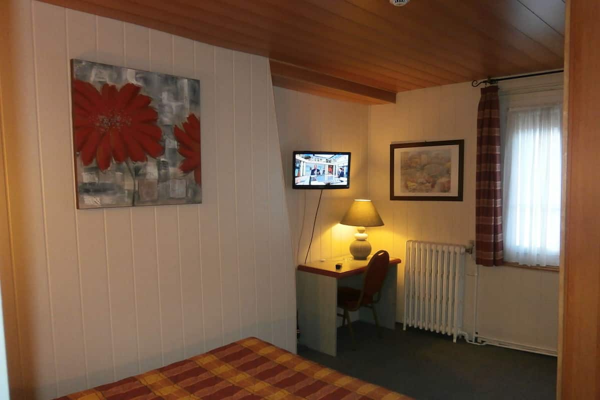 Quarto com cama, escrivaninha com luminária, cadeira, radiador e TV na parede.