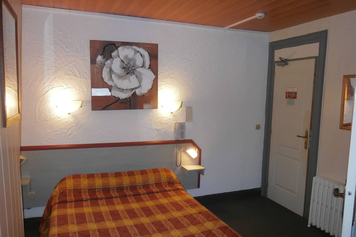 Quarto com cama, luminária na parede, quadro de uma flor e uma porta.