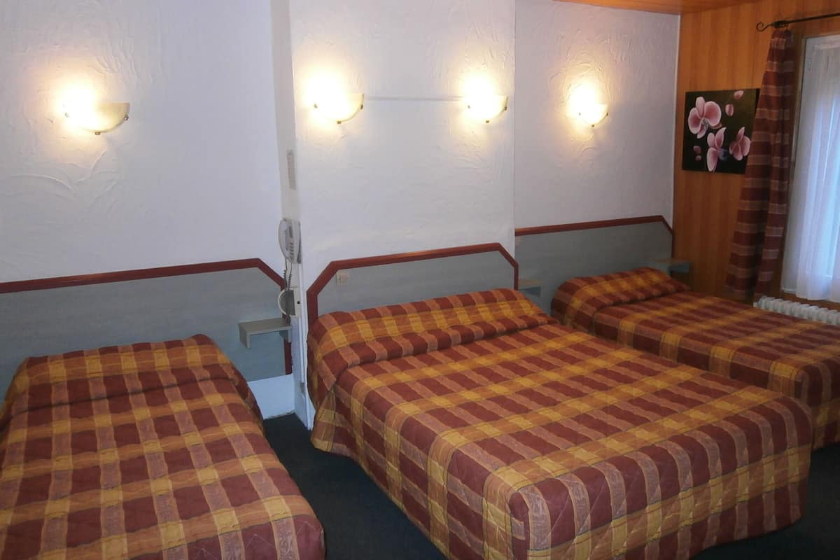 Quarto de hotel com três camas, cada uma com colcha xadrez. Há luminárias fixadas na parede sobre as camas, e um telefone na parede.