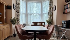 Uma área de refeições com uma mesa redonda, quatro cadeiras, um candelabro, uma estante e vista para árvores através de grandes janelas.