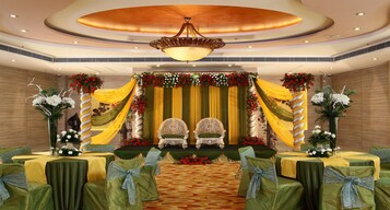 Um palco com duas cadeiras ornamentadas, cobertas com tecido amarelo e verde, e rodeado por arranjos florais. Mesas com toalhas verdes e fitas azuis estão dispostas em primeiro plano.