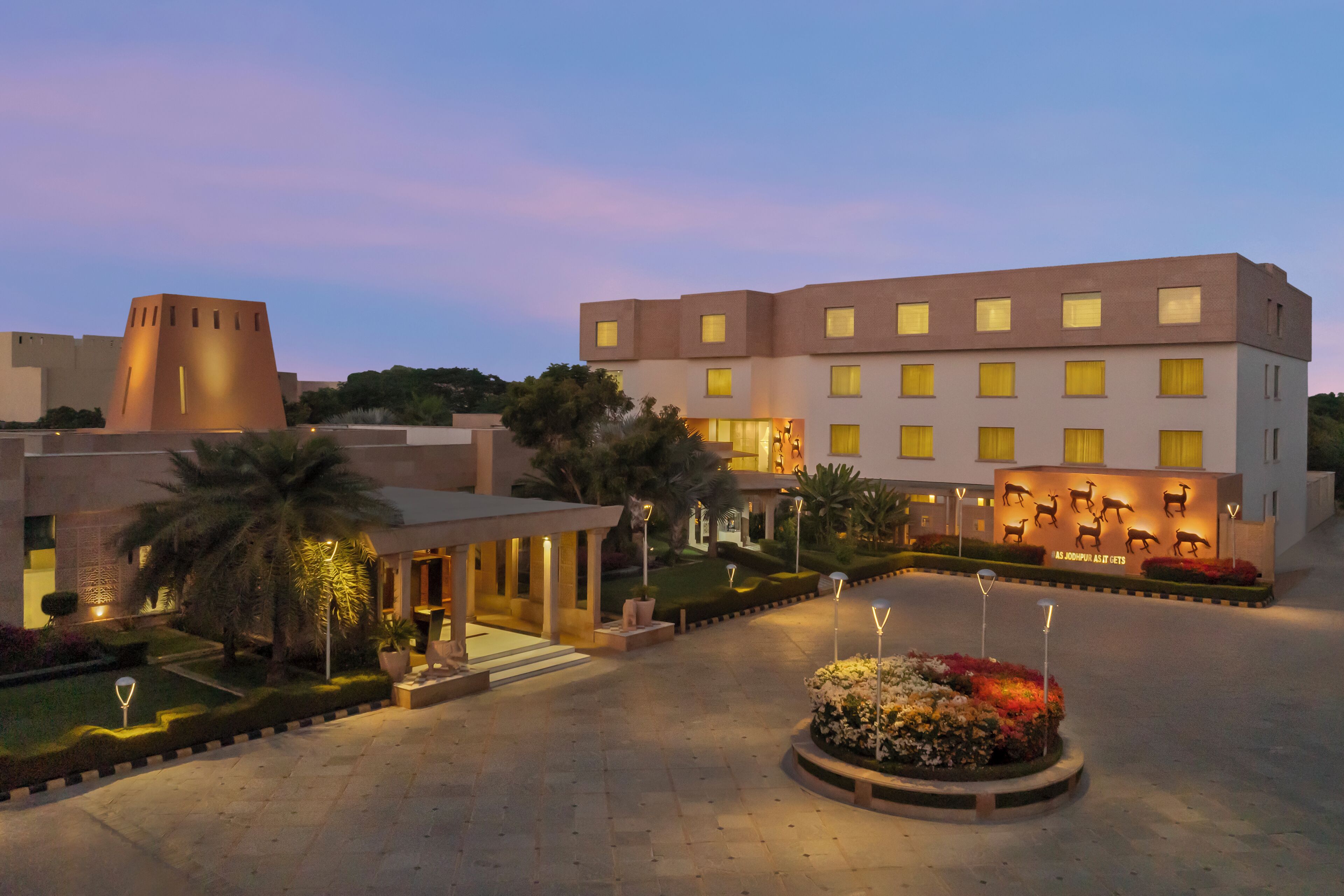 Hotel com design moderno, apresentando um pátio com arranjo floral central, área de estar coberta e um edifício imponente com várias janelas e estrutura em torre.
