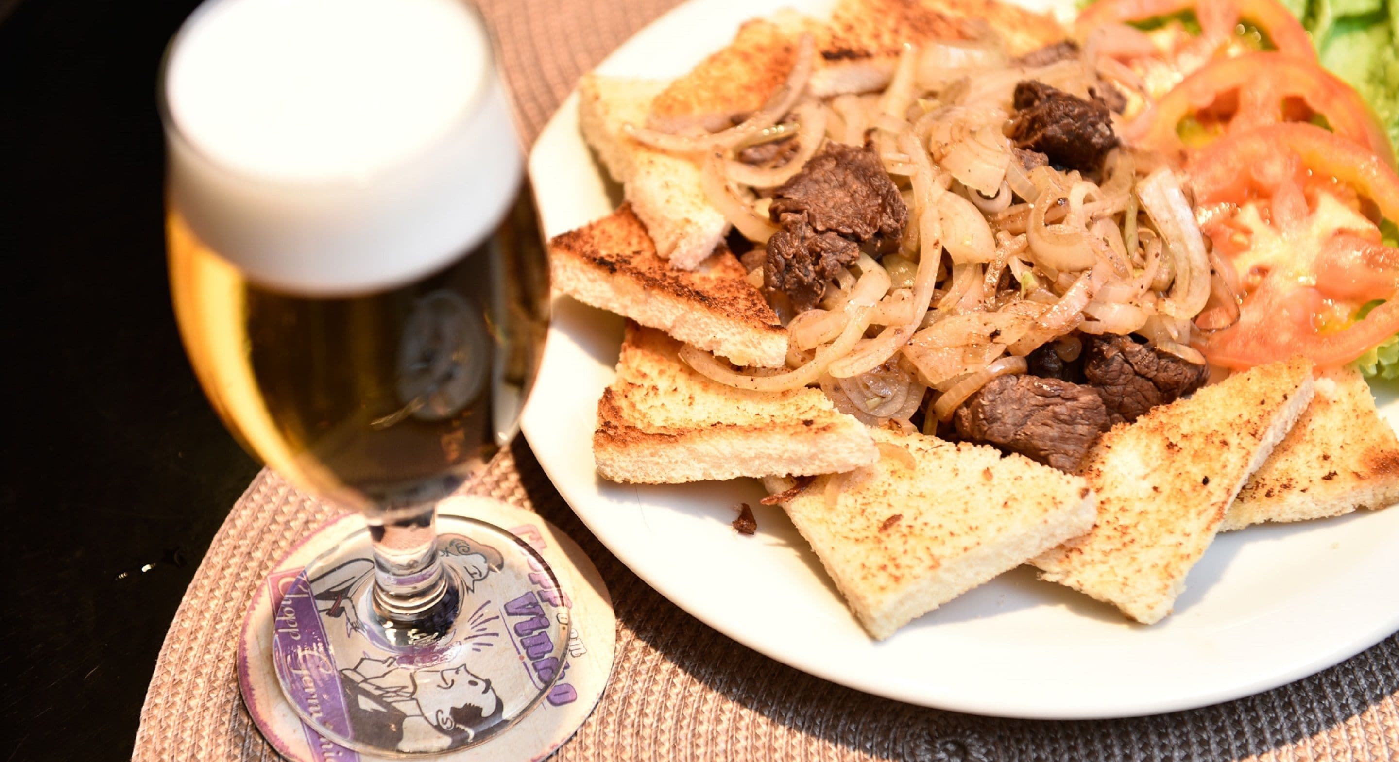 Prato de comida com pão, cebolas e carne, e um copo de cerveja sobre uma mesa.