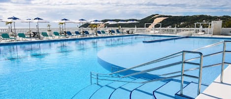 Piscina azul límpida com espreguiçadeiras e guarda-sóis, cercada por um muro branco e vista para colinas.