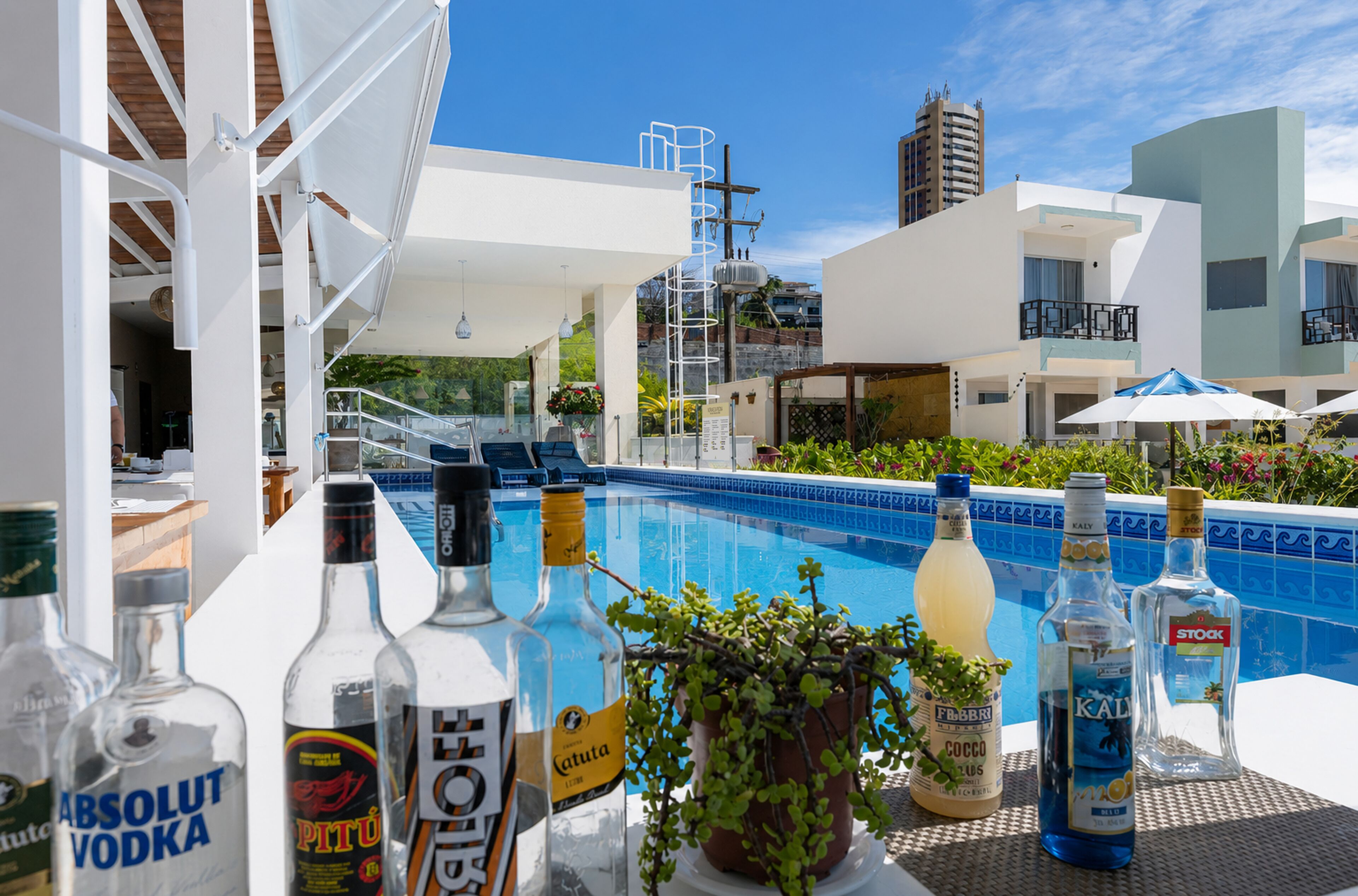 Área de piscina com diversos tipos de bebidas alcoólicas, uma piscina e edifícios modernos ao fundo.