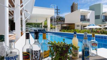 Área de piscina com diversos tipos de bebidas alcoólicas, uma piscina e edifícios modernos ao fundo.