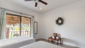 Quarto de hotel com duas camas, um ventilador de teto, uma mesinha com cesto e toalhas, uma guirlanda na parede e vista para o exterior por uma grande janela.