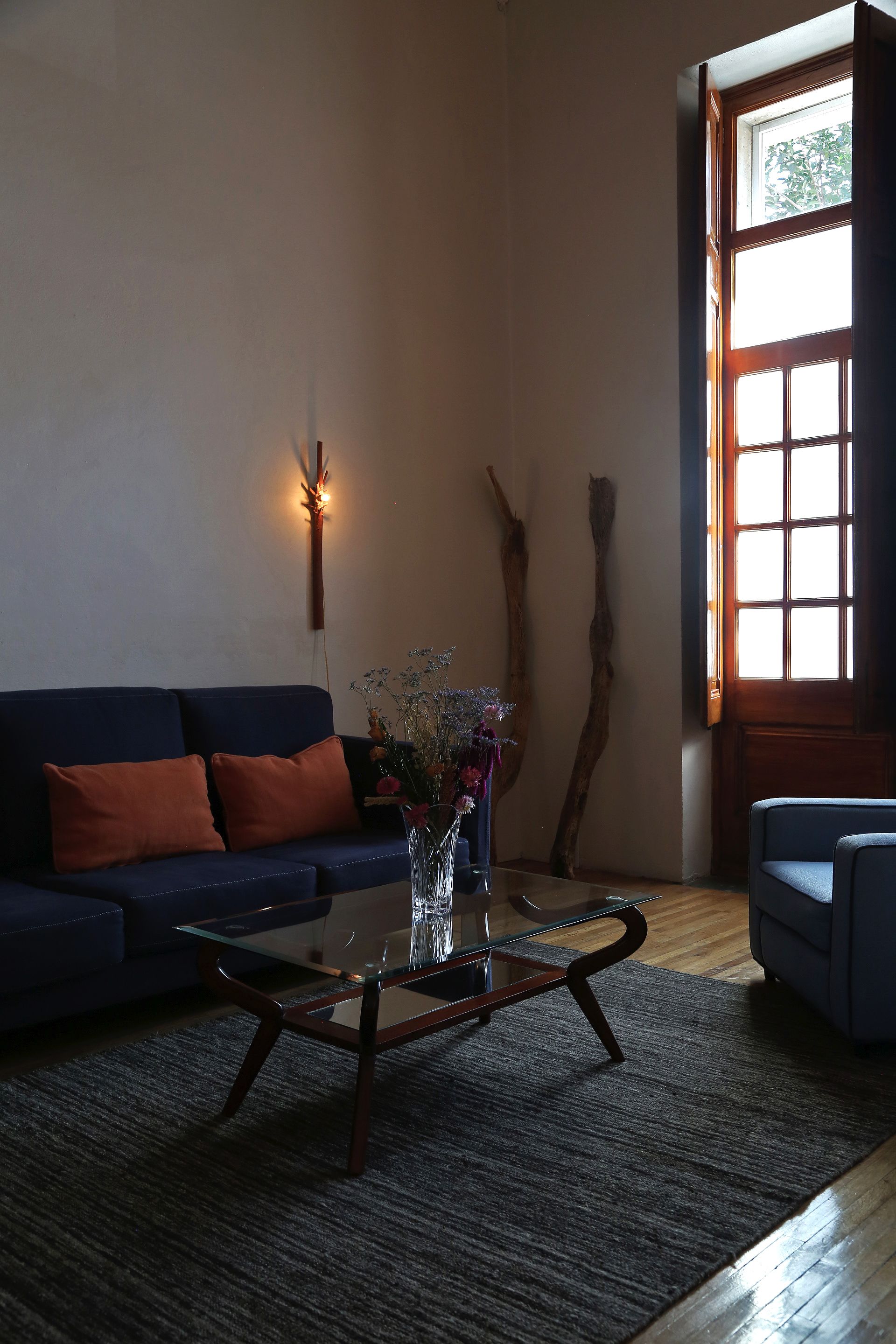 Uma sala de estar com um sofá azul escuro, almofadas laranja, uma mesa de centro de vidro e um vaso com flores. Há uma escultura de madeira na parede e uma janela com luz natural.