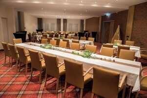 Uma sala de conferências com mesas compridas e cadeiras, um tapete xadrez vermelho e paredes de tijolo.