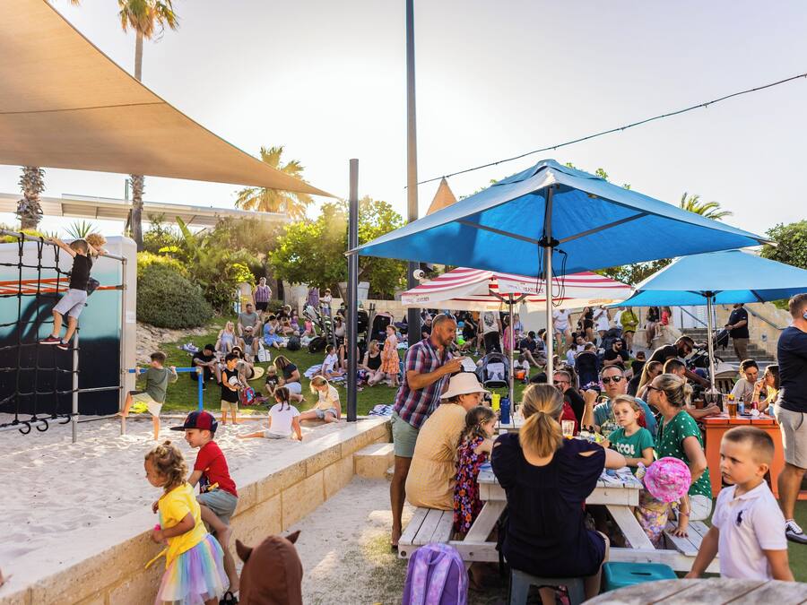 Um evento ao ar livre movimentado, com pessoas sentadas em mesas sob guarda-sóis, uma área de recreação infantil com estrutura de escalada e uma área arenosa.