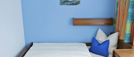 Uma cama bem arrumada com roupa de cama branca e um travesseiro azul, cabeceira de madeira e uma pequena pintura de uma gota d'água na parede.