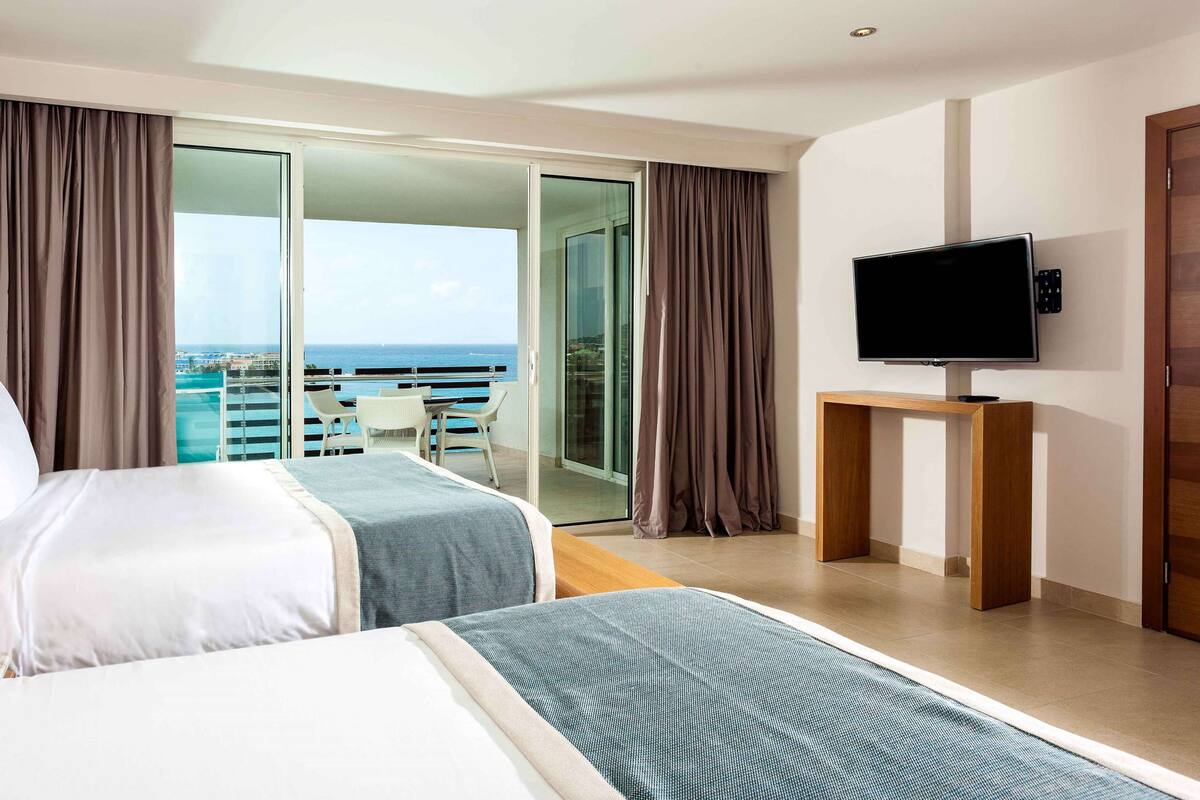 Quarto de hotel com duas camas, televisão e varanda com vista para o mar.