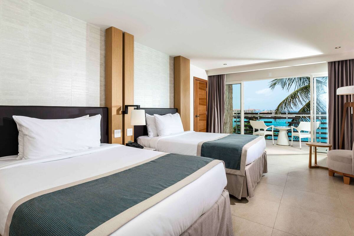Quarto de hotel com duas camas, varanda com piscina e vista para o mar.