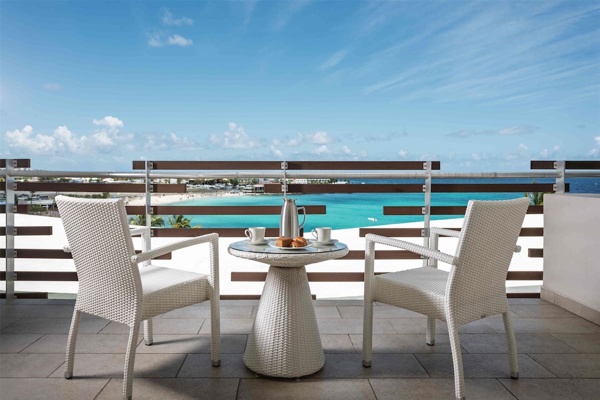Varanda com cadeiras de vime e uma mesa, com vista para uma praia e águas turquesa.