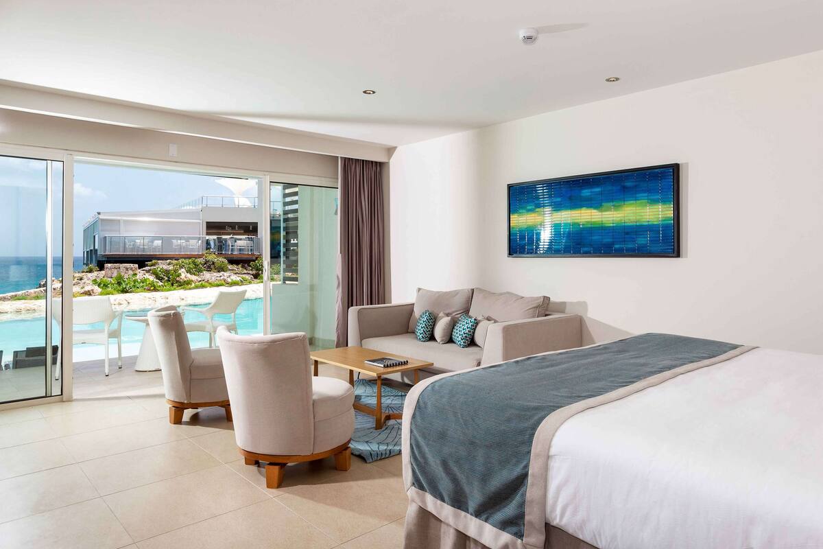 Quarto de hotel com cama, sofá, mesa de centro e mesa de jantar. Há vista para o oceano e uma piscina fora do quarto.