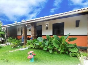 Uma casa com telhado de telhas, varanda frontal e um jardim bem cuidado com plantas grandes e folhosas e uma decoração de cogumelo.