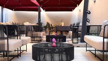Uma área de estar com mesas e cadeiras redondas, um vaso decorativo central e um guarda-sol vermelho acima.