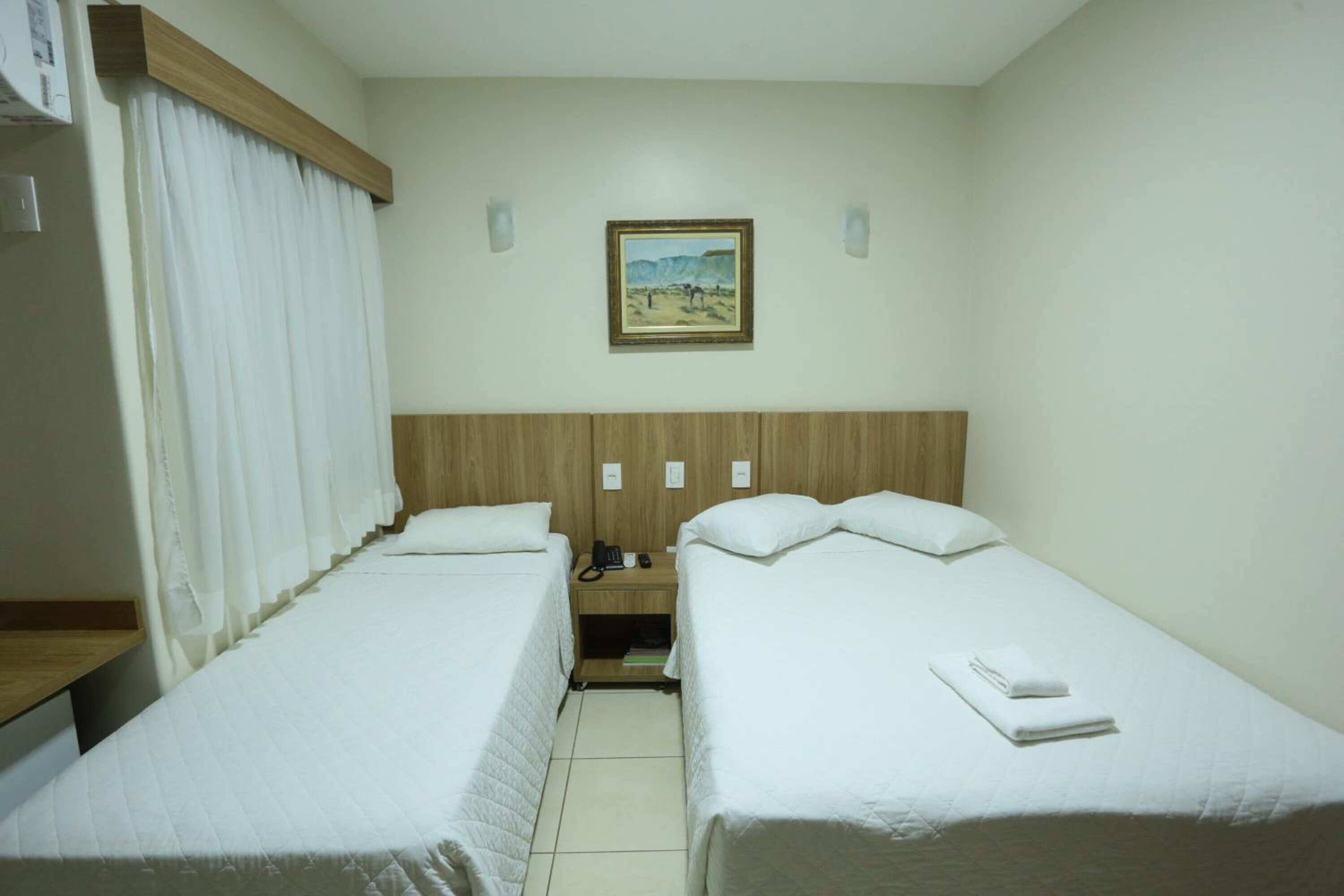 Quarto de hotel com duas camas de solteiro, cabeceira de madeira, mesa de cabeceira com telefone e um quadro na parede.