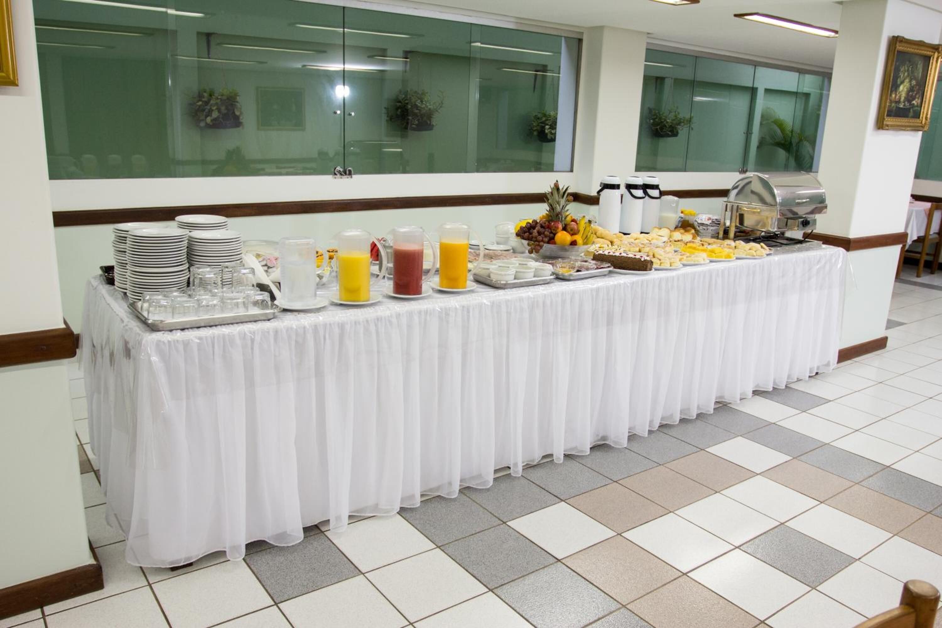 Mesa de buffet com diversos alimentos e bebidas, cercada por janelas de vidro e piso ladrilhado.