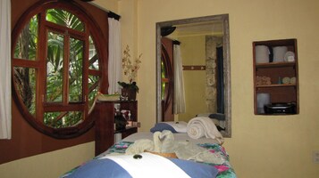 Quarto com cama, espelho, janela com cortinas, prateleira com toalhas e uma pequena planta.