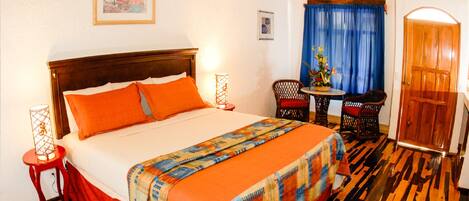 Quarto de hotel com piso de madeira, cama com roupa de cama laranja, cabeceira de madeira, mesa pequena com cadeiras e uma porta.