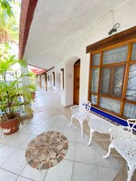 Uma área externa coberta com piso de azulejos, cadeiras de metal branco e plantas em vasos.