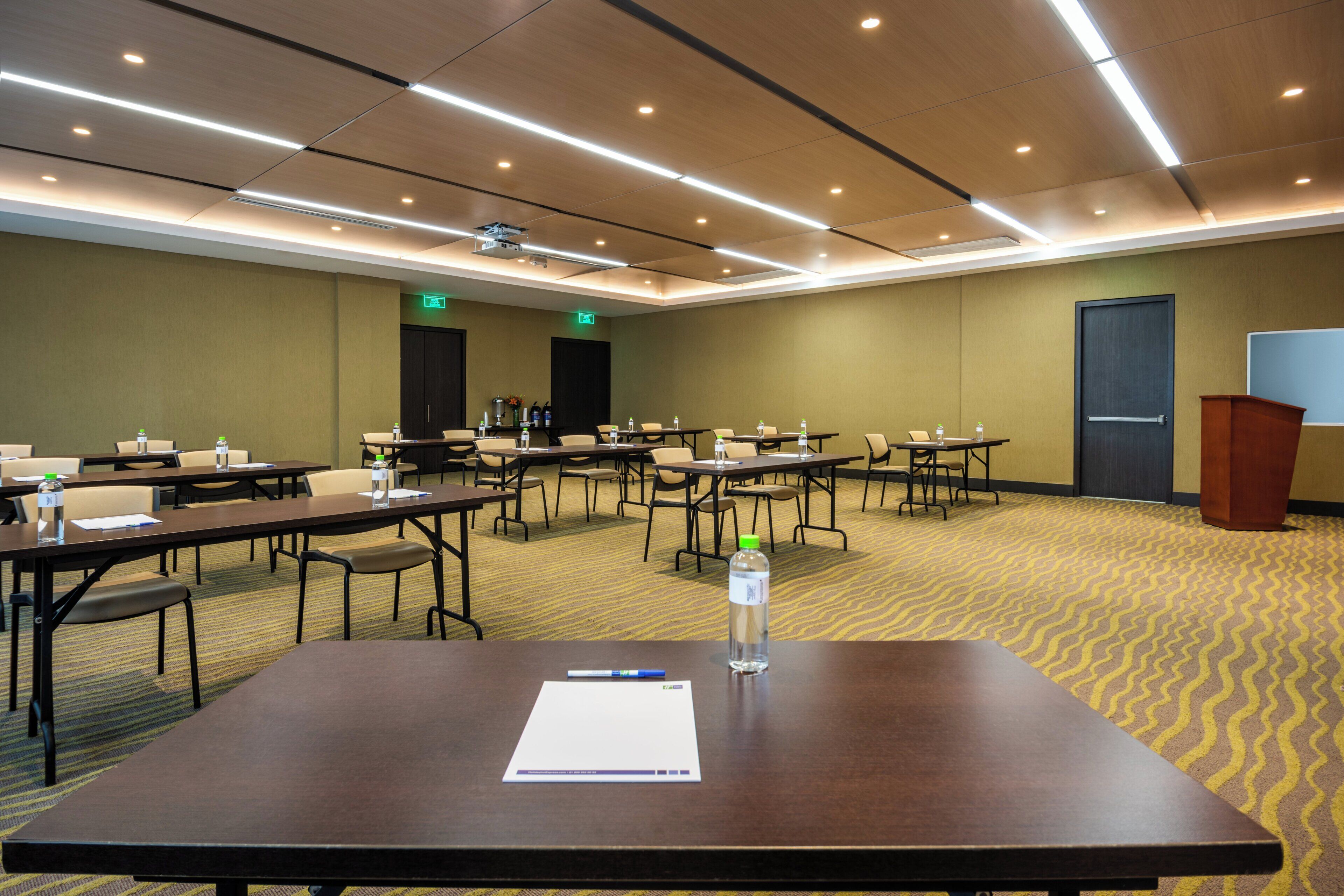 Uma sala de conferências com mesas, cadeiras, um pódio e uma tela de projeção.
