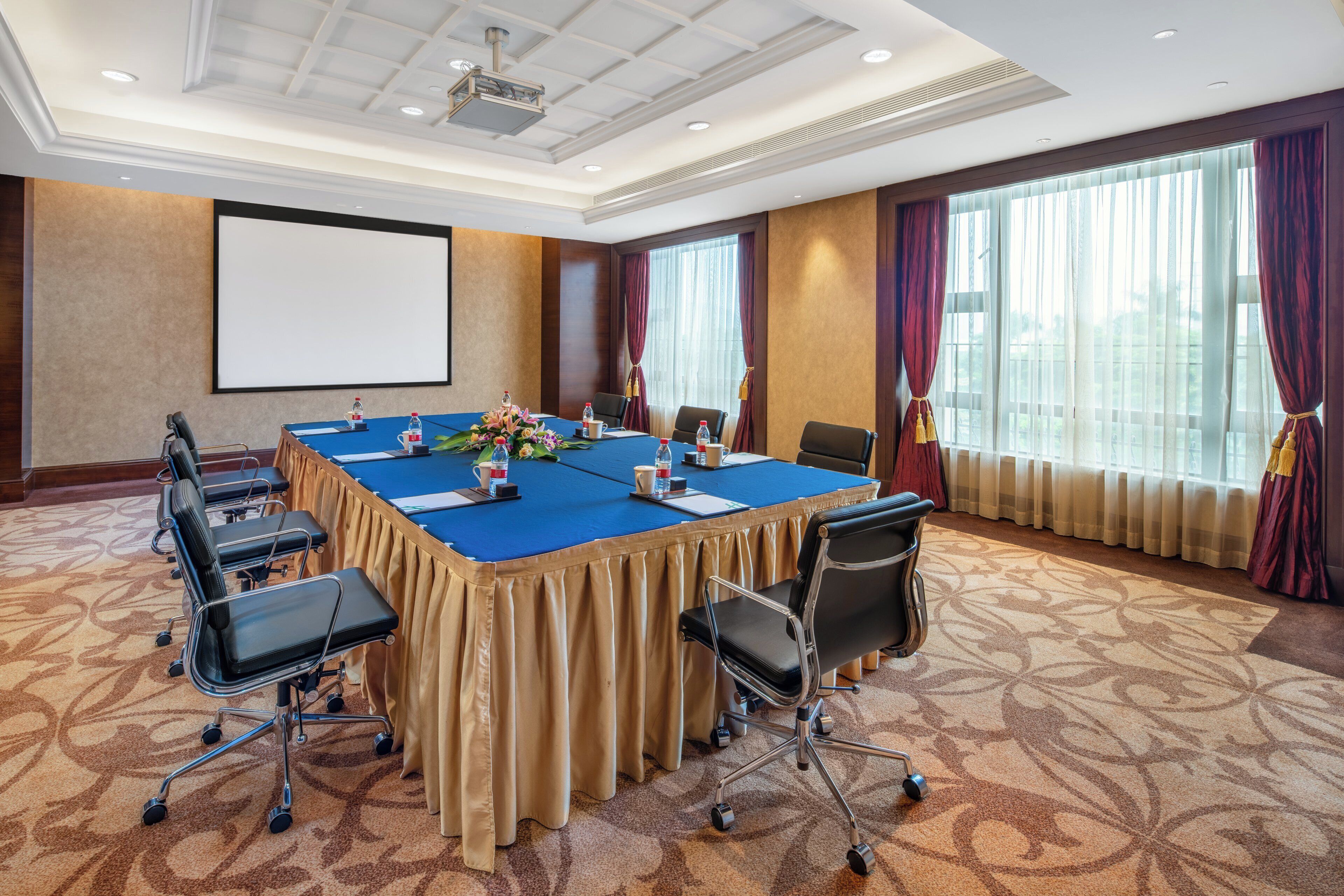 Sala de conferências com uma mesa comprida, cadeiras, tela de projetor e uma grande janela com cortinas.
