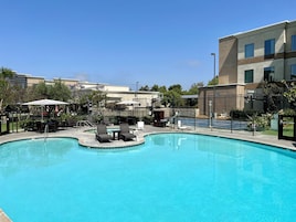 Piscina com espreguiçadeiras e guarda-sóis, cercada por um muro e edifícios.