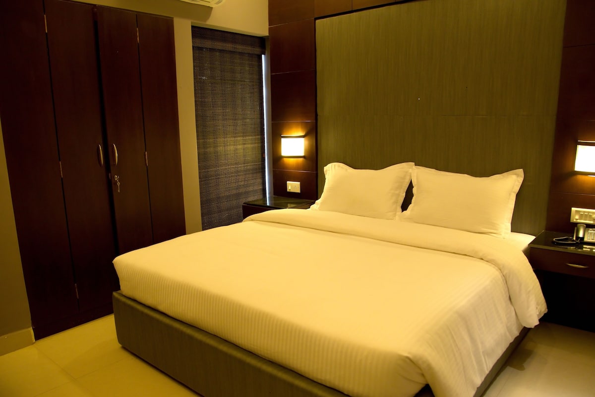 Quarto de hotel com uma cama grande, um guarda-roupa, uma mesa de cabeceira com luminária e uma luz na parede.