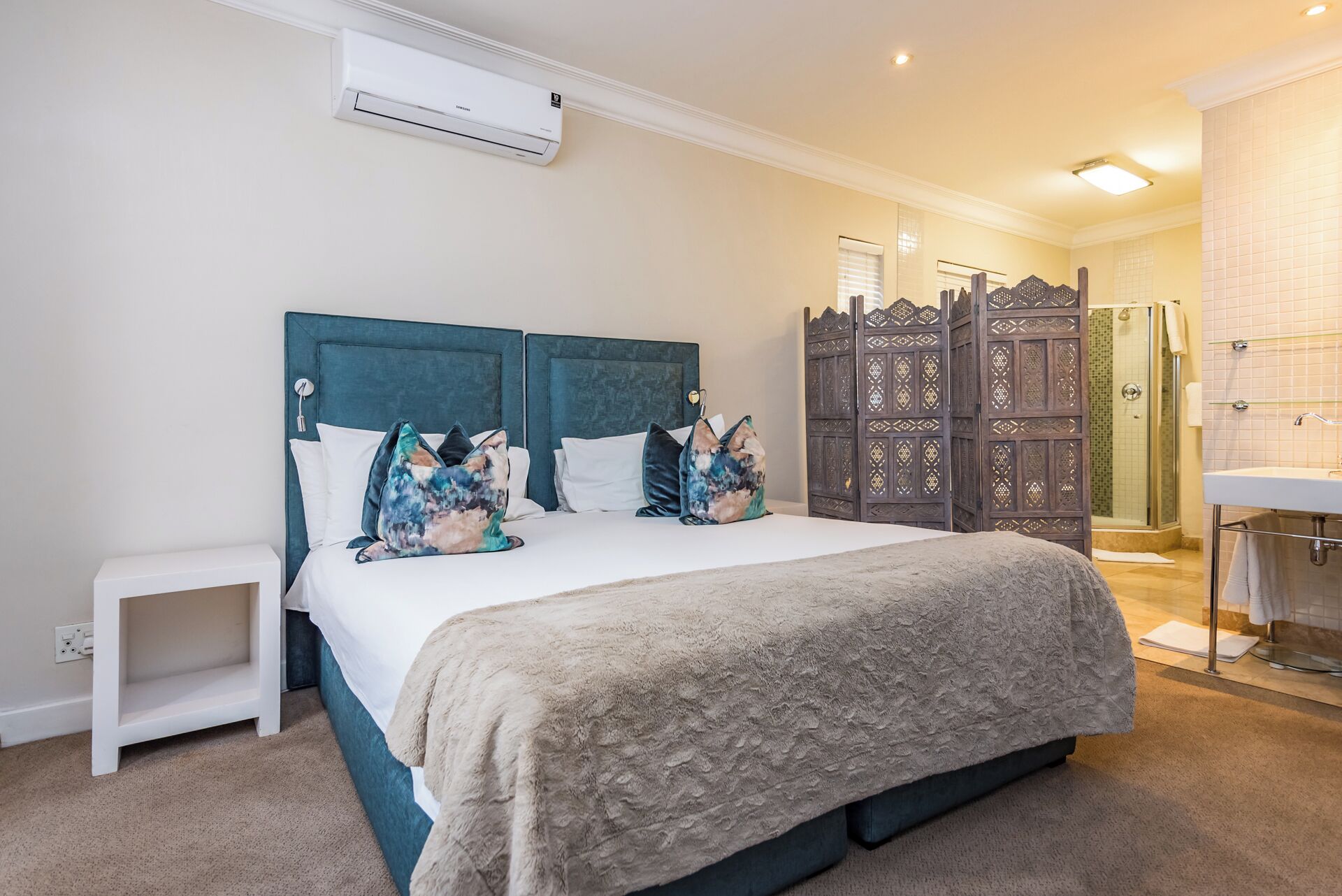 Quarto com cama, mesa de cabeceira, aparelho de ar condicionado, chuveiro e banheiro com pia.