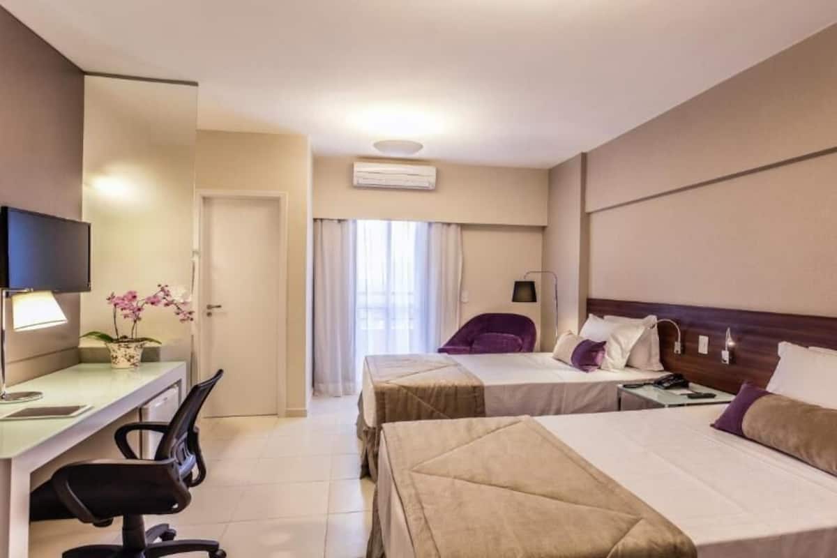 Quarto de hotel com duas camas, uma escrivaninha com cadeira, televisão e janela com cortinas.