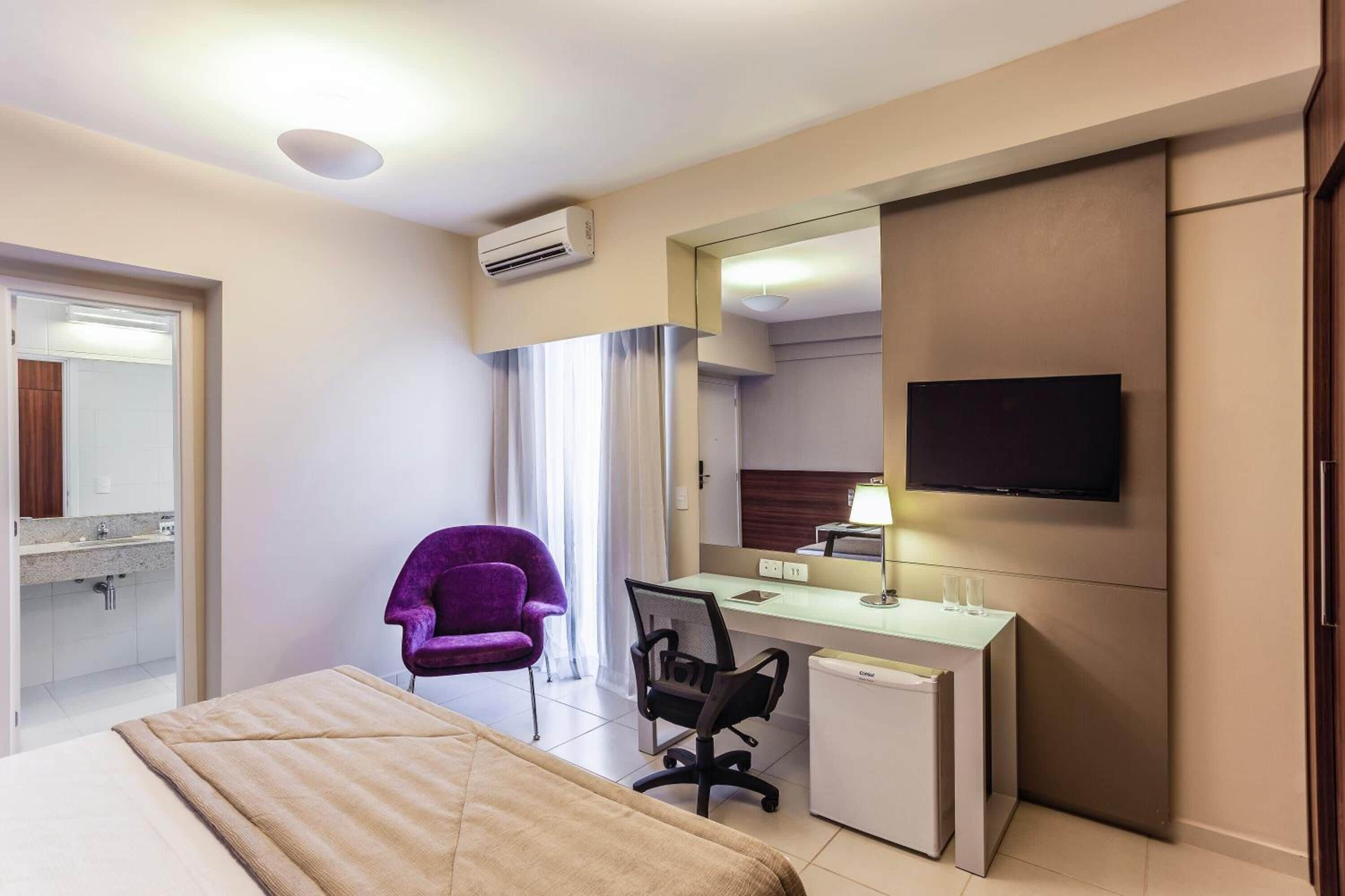Quarto de hotel com cama, escrivaninha, cadeira, televisão e ar condicionado.