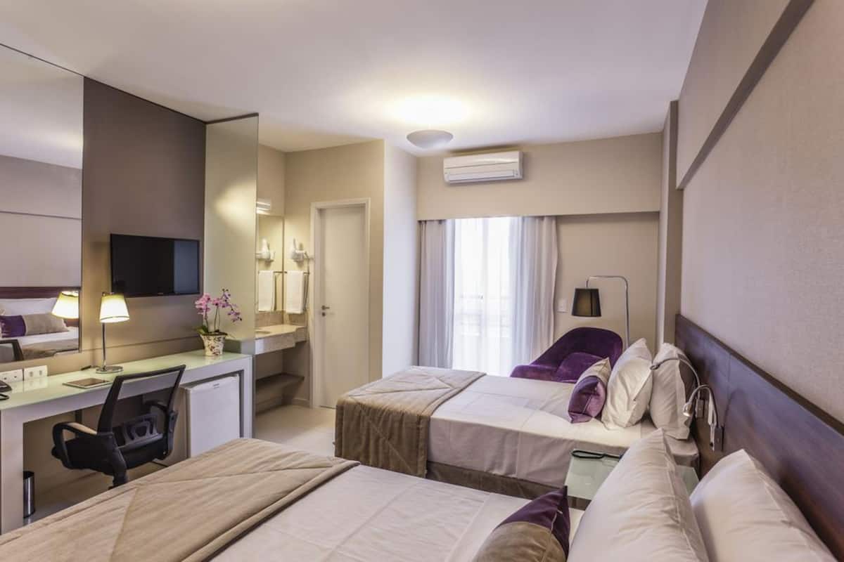 Quarto de hotel com duas camas, uma escrivaninha, uma cadeira, uma televisão e um ar condicionado.