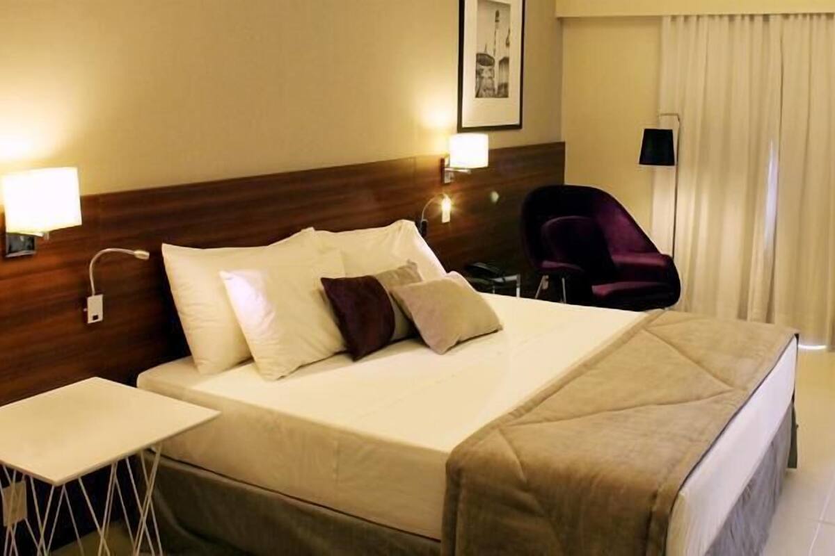 Quarto de hotel com cama, mesa de cabeceira, cadeira e luminária na parede.