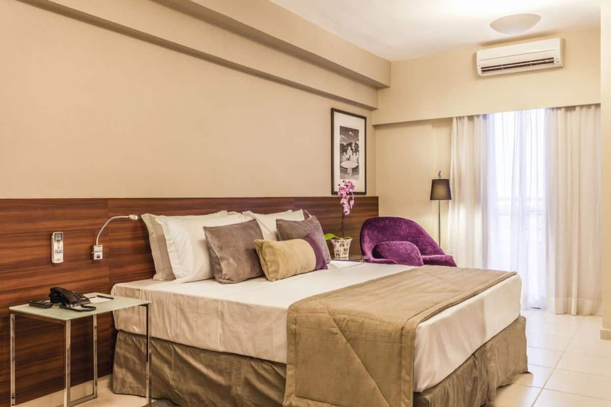 Quarto de hotel com cama, mesa de cabeceira, telefone, ar condicionado e uma cadeira.